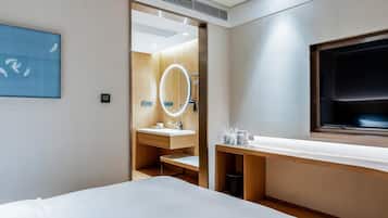 Quarto de hotel com cama, espelho grande iluminado, televisão e banheiro com pia e chuveiro.