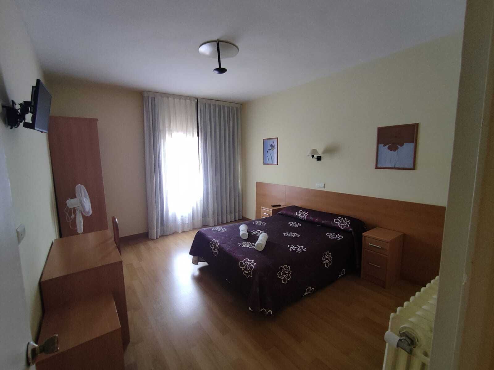 Quarto de hotel com cama de madeira, uma escrivaninha, cadeira, radiador e janela com cortinas.