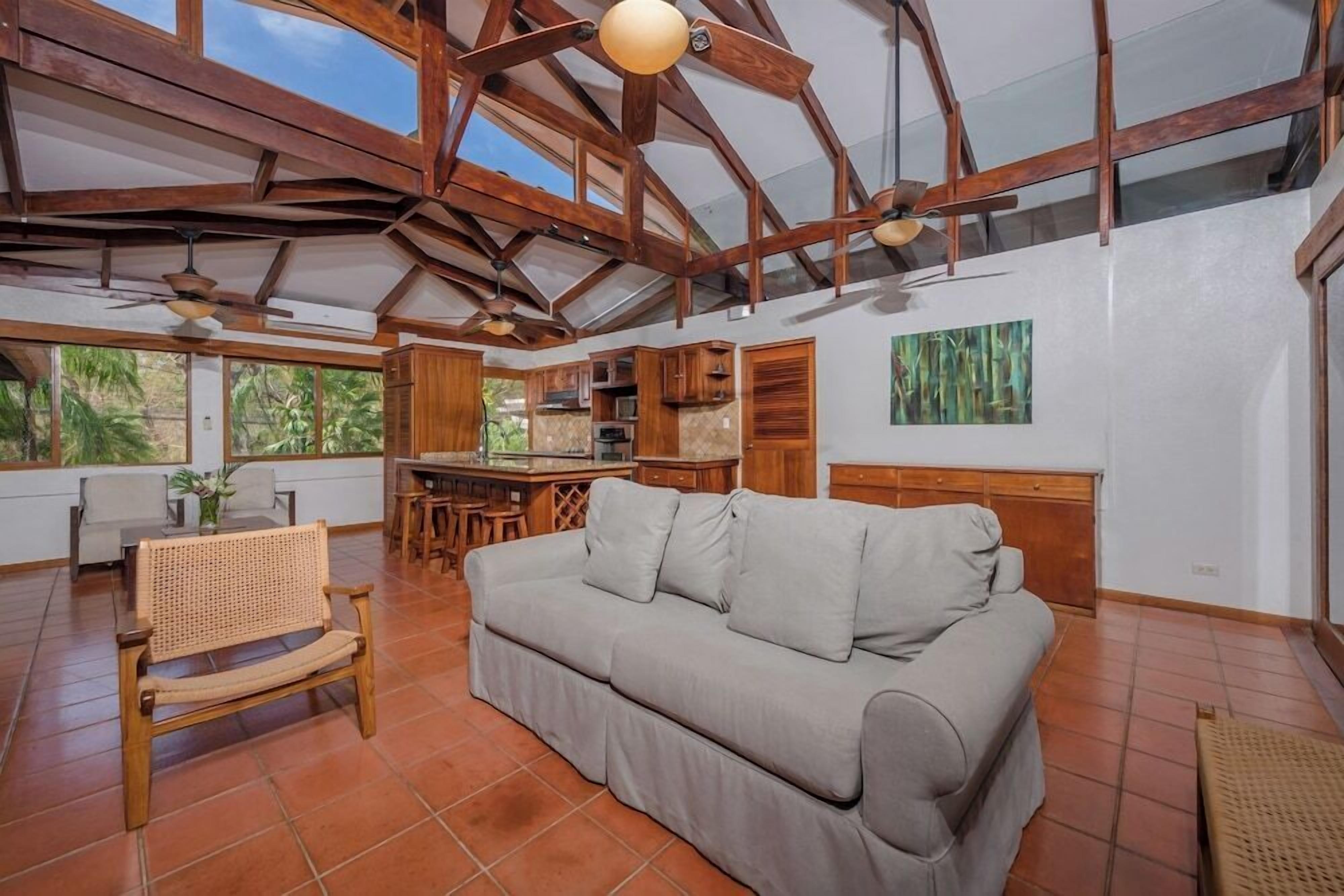 Ampla sala de estar com teto de madeira, um sofá, um banco e uma área de jantar com cozinha ao fundo.