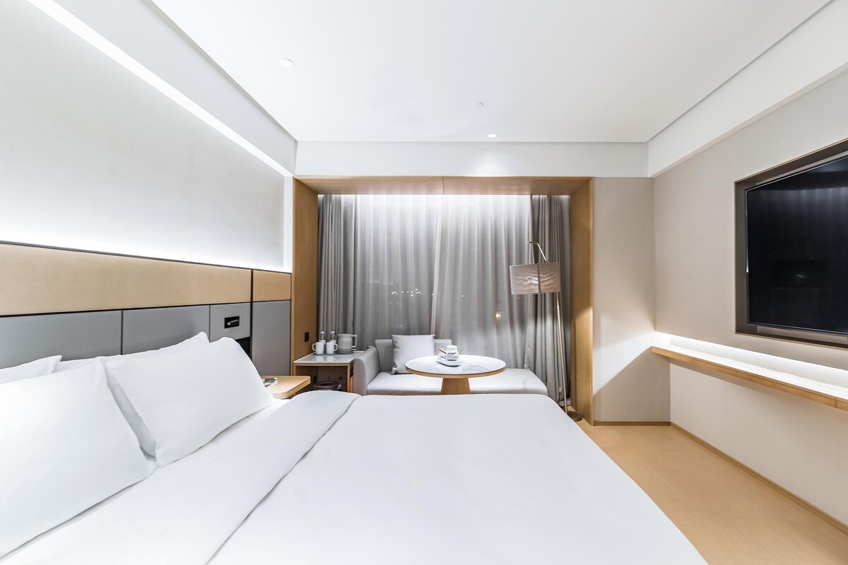 Quarto de hotel moderno com uma cama grande, mesas de cabeceira, uma mesinha redonda e uma janela.