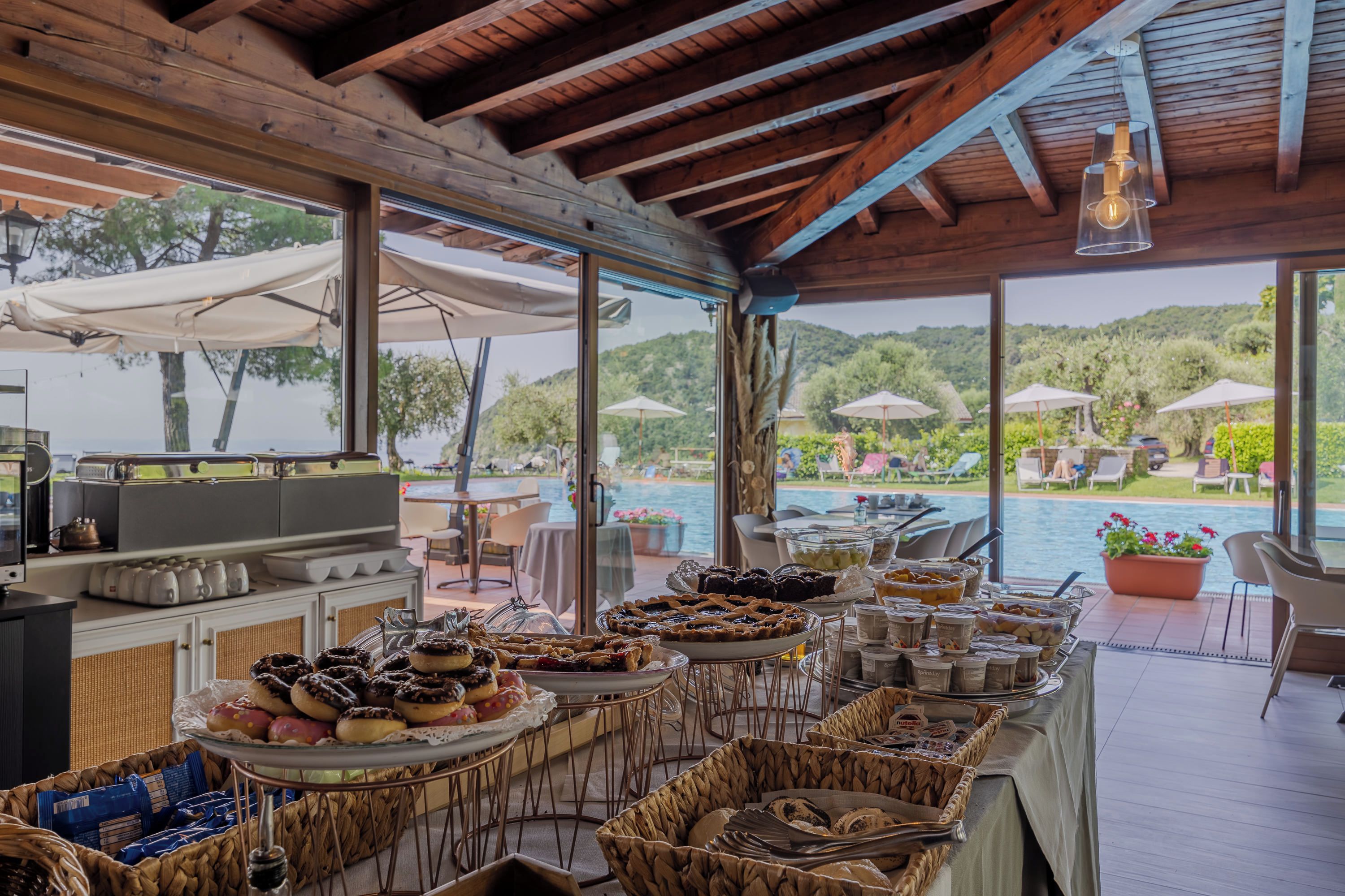 Um buffet de café da manhã com diversos pães e bebidas, com vista para uma piscina e área de jardim.
