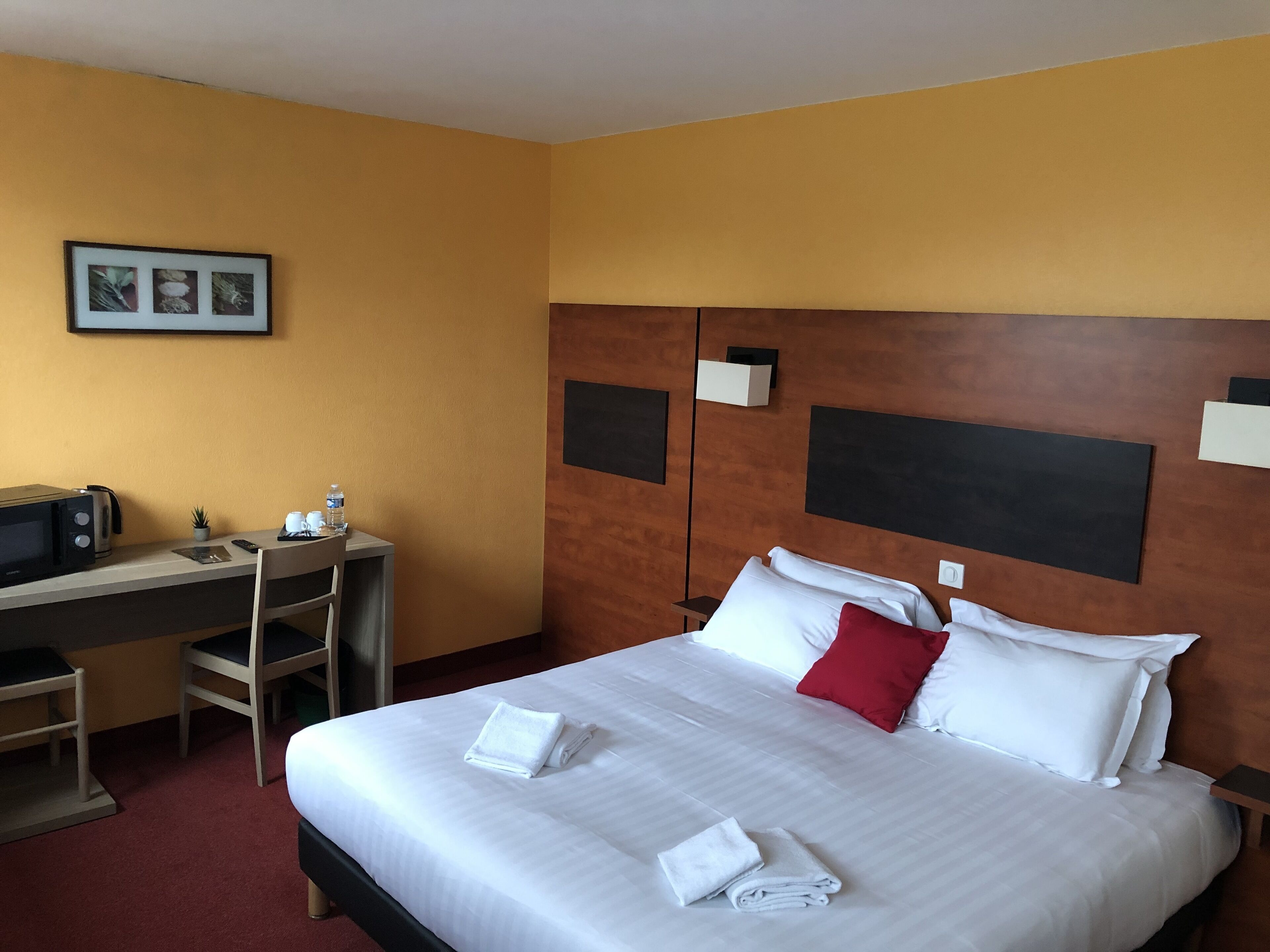 Quarto de hotel com cama, escrivaninha com micro-ondas, cadeira e pequena mesa com garrafa de água e copos.