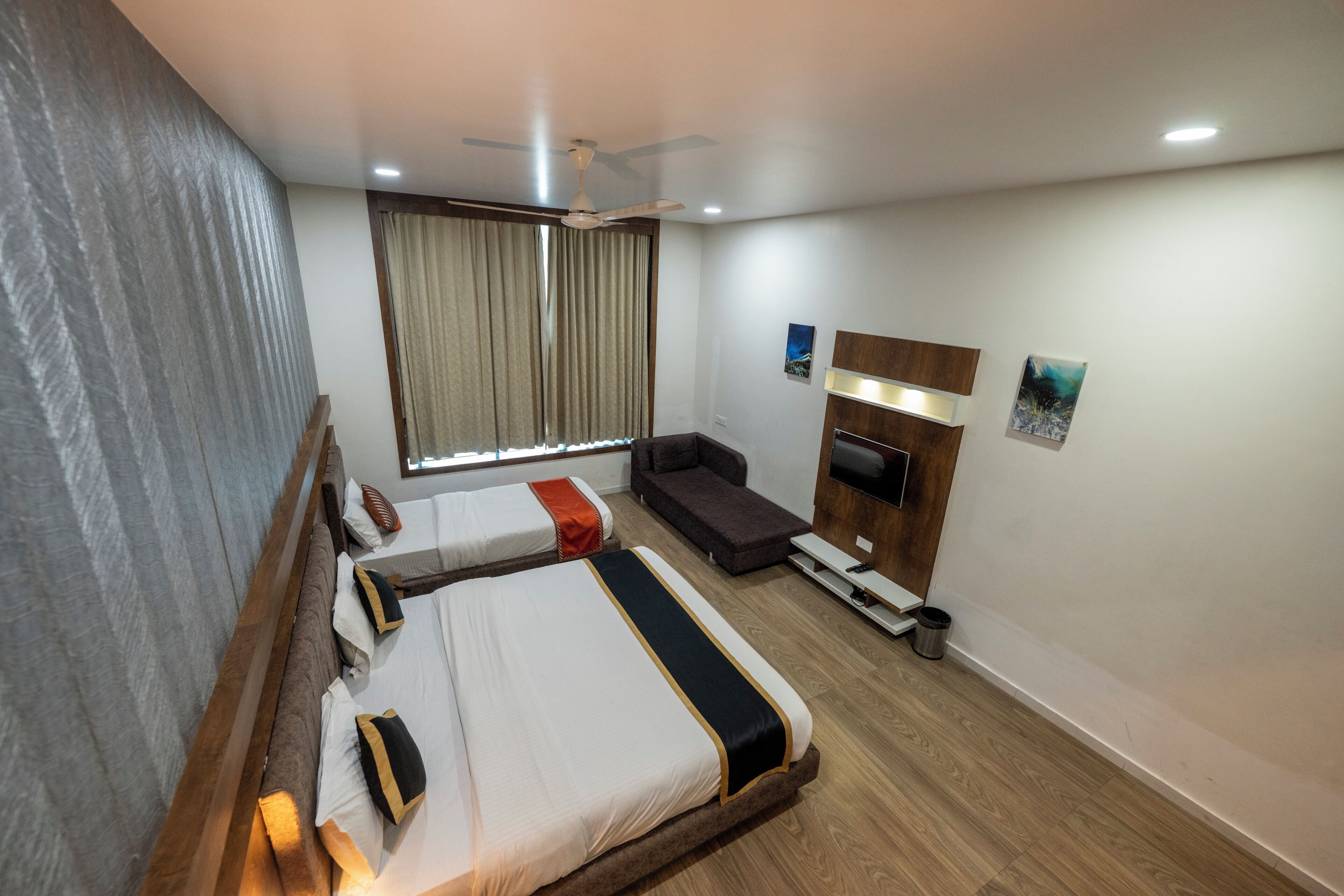 Quarto de hotel com duas camas, um sofá, uma televisão, um ventilador de teto e uma janela com cortinas.