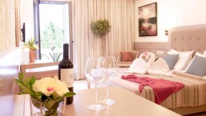 Quarto de hotel com uma cama, uma mesa com vinho e copos, um vaso com flores e vista para o exterior.