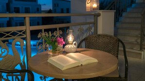 Mesa redonda com um livro aberto, uma vela e um vaso de flores numa varanda com vista para um edifício.
