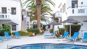 Uma área de piscina com espreguiçadeiras azuis e guarda-sóis, cercada por edifícios brancos com varandas.