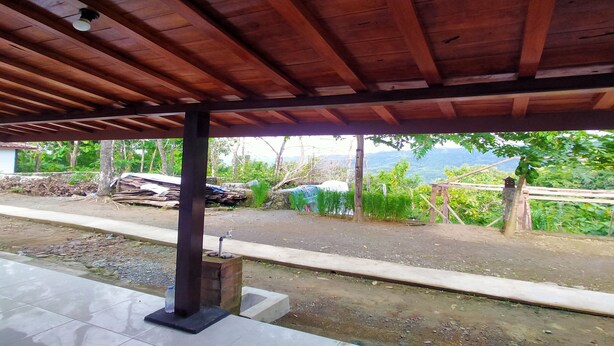 Um espaço ao ar livre coberto, com teto de madeira, um banco de concreto e vista para uma paisagem rural com árvores e um caminho de terra.