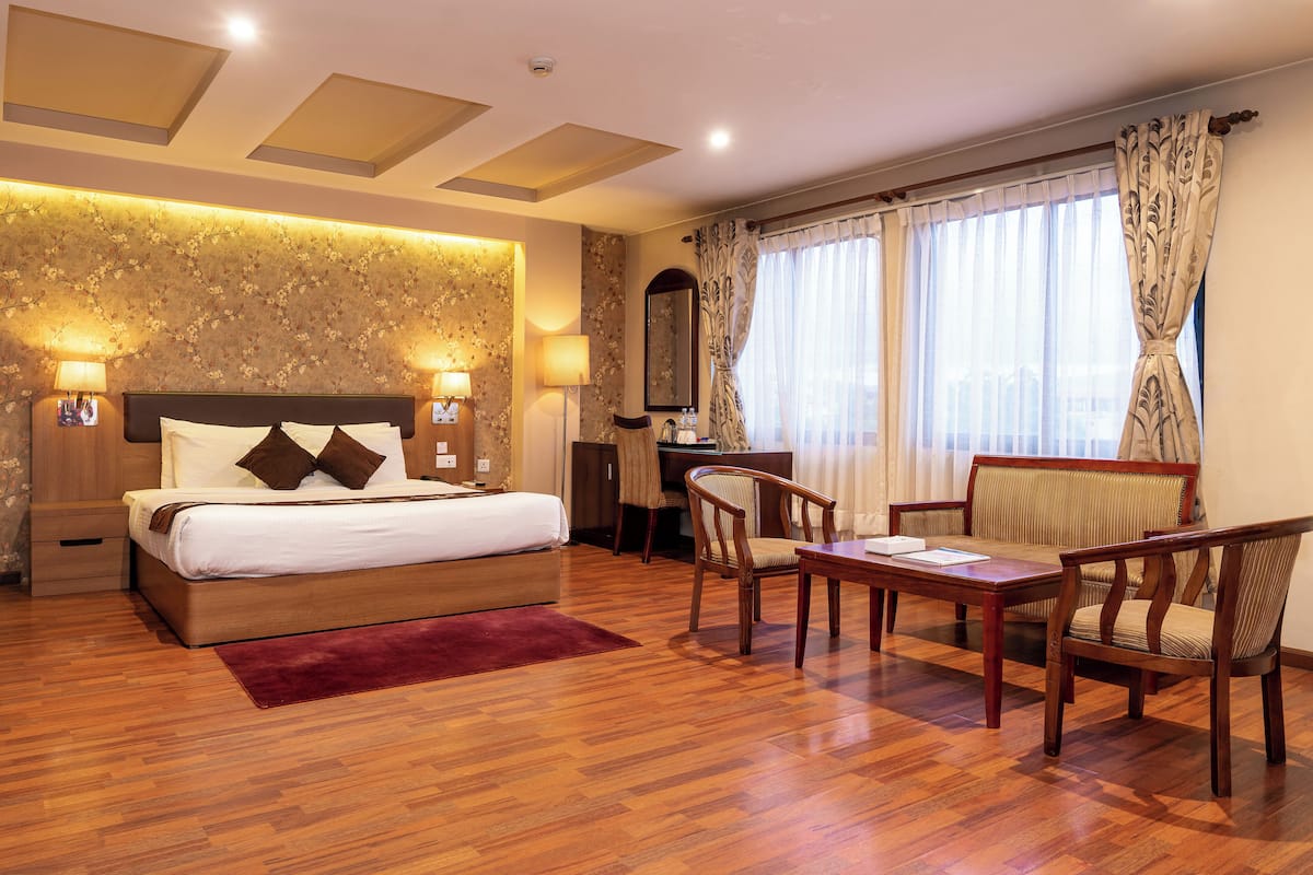 Quarto de hotel com uma cama grande, uma escrivaninha, duas cadeiras e uma janela ampla com cortinas.