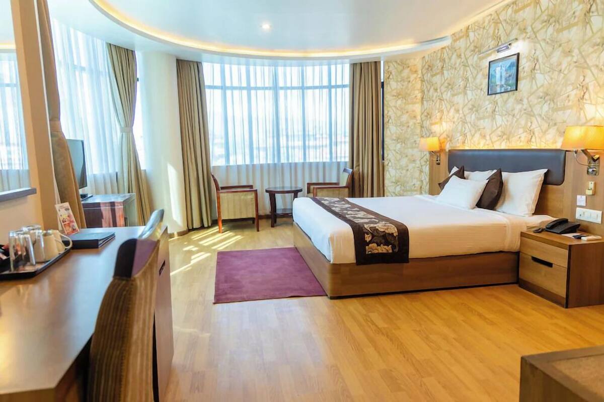 Quarto de hotel com cama, mesa de cabeceira, luminária e escrivaninha com cadeira.