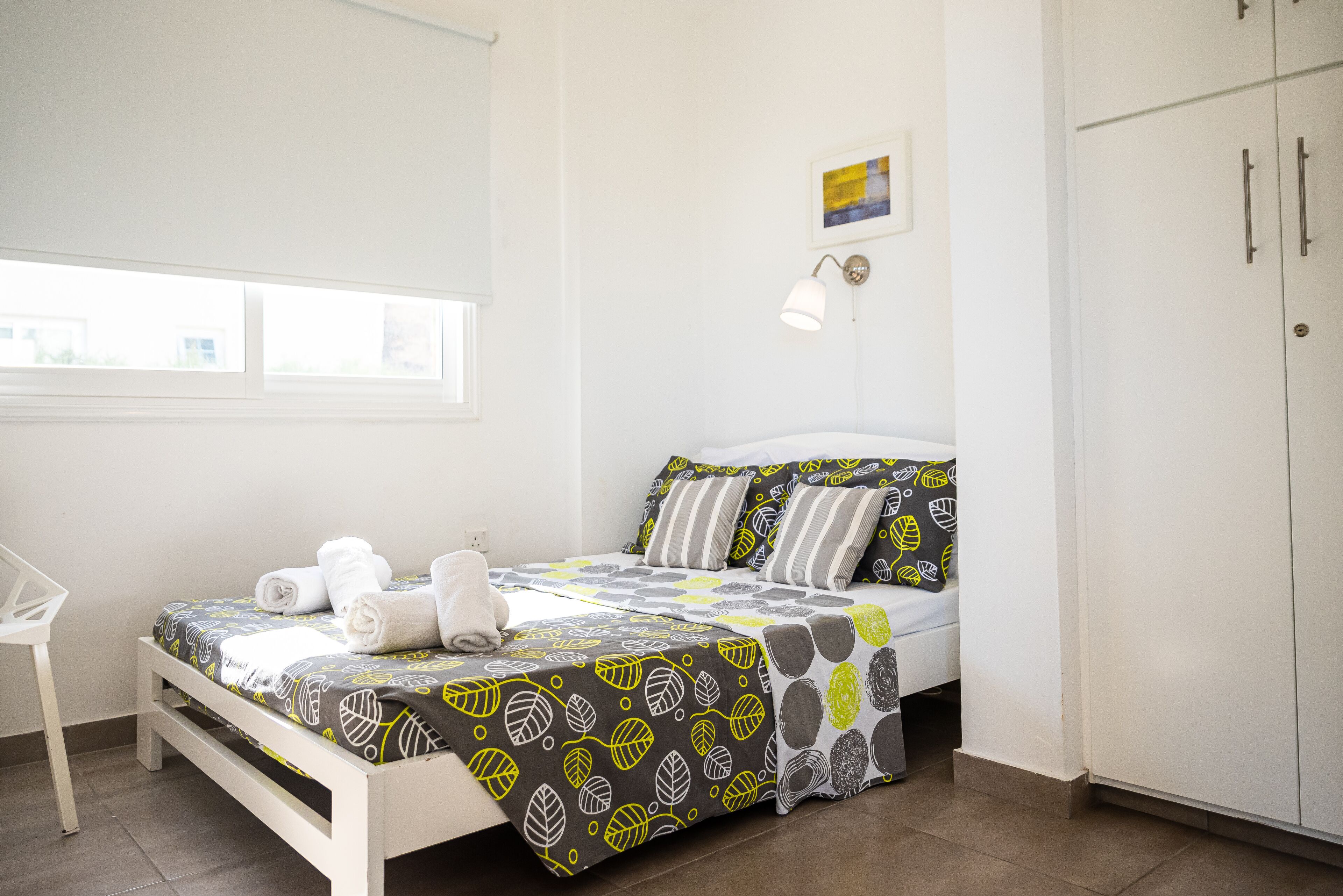 Uma cama bem arrumada com colcha estampada, um abajur e uma janela com persianas.