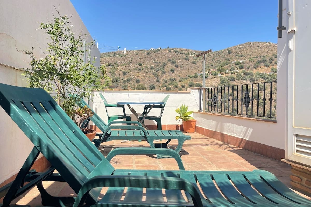 Terraço com cadeiras de descanso verdes e uma mesa, com vista para uma paisagem montanhosa.