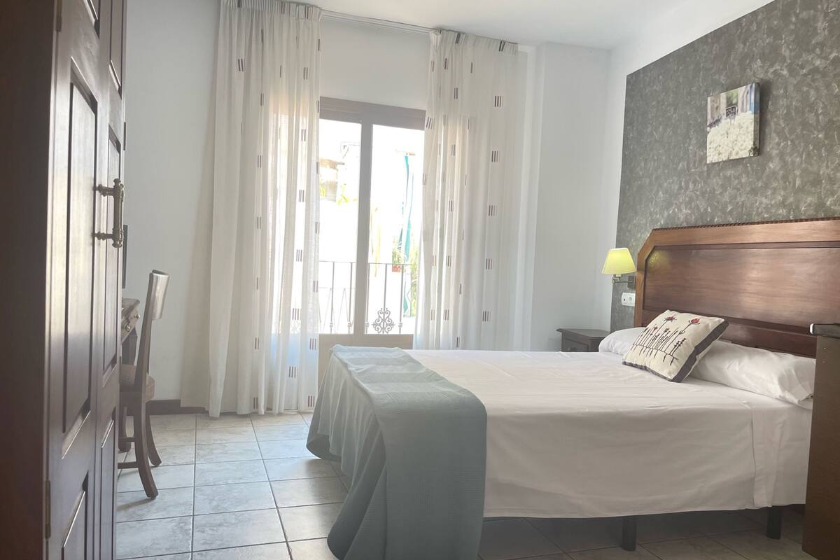 Quarto de hotel com cama, mesa de cabeceira, luminária, janela com cortinas e porta.