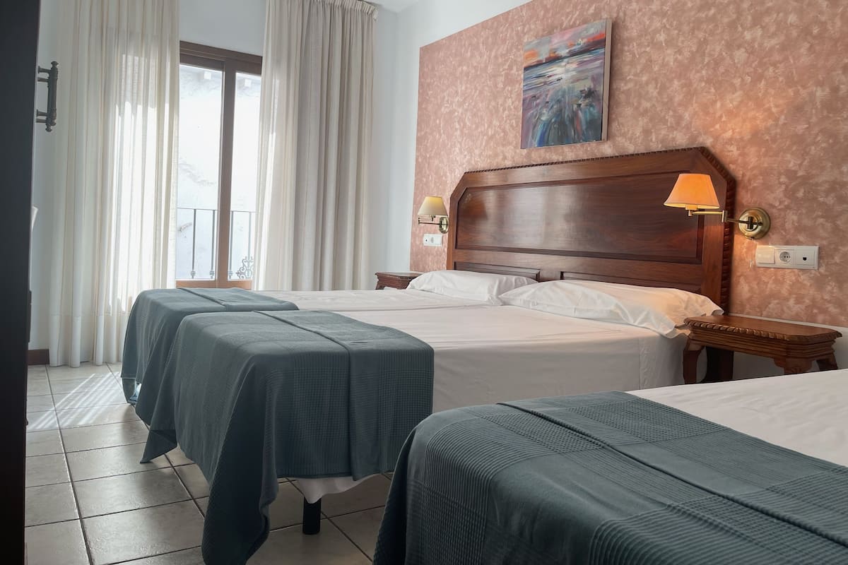 Quarto de hotel com duas camas, uma janela grande com cortinas, um quadro na parede e uma mesa de cabeceira com abajur.
