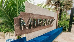 Um letreiro com a palavra 'MIRRAGE' em letras grandes, fixado em uma estrutura de pedra com base azul, rodeado por vegetação.