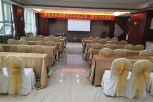 Uma sala de conferências com fileiras de mesas e cadeiras, uma tela de projeção e um banner na parede.