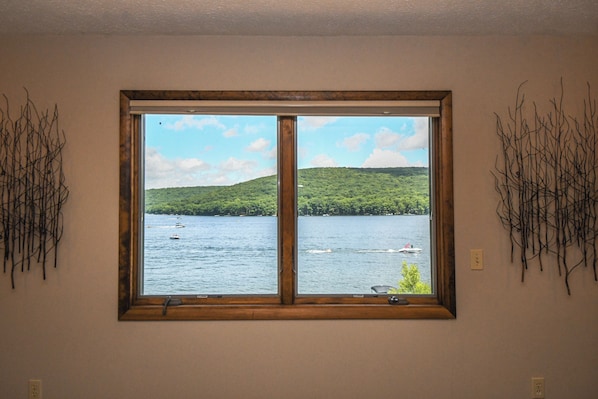 Quarto com uma grande janela que oferece vista para um lago com barcos e uma colina verde.