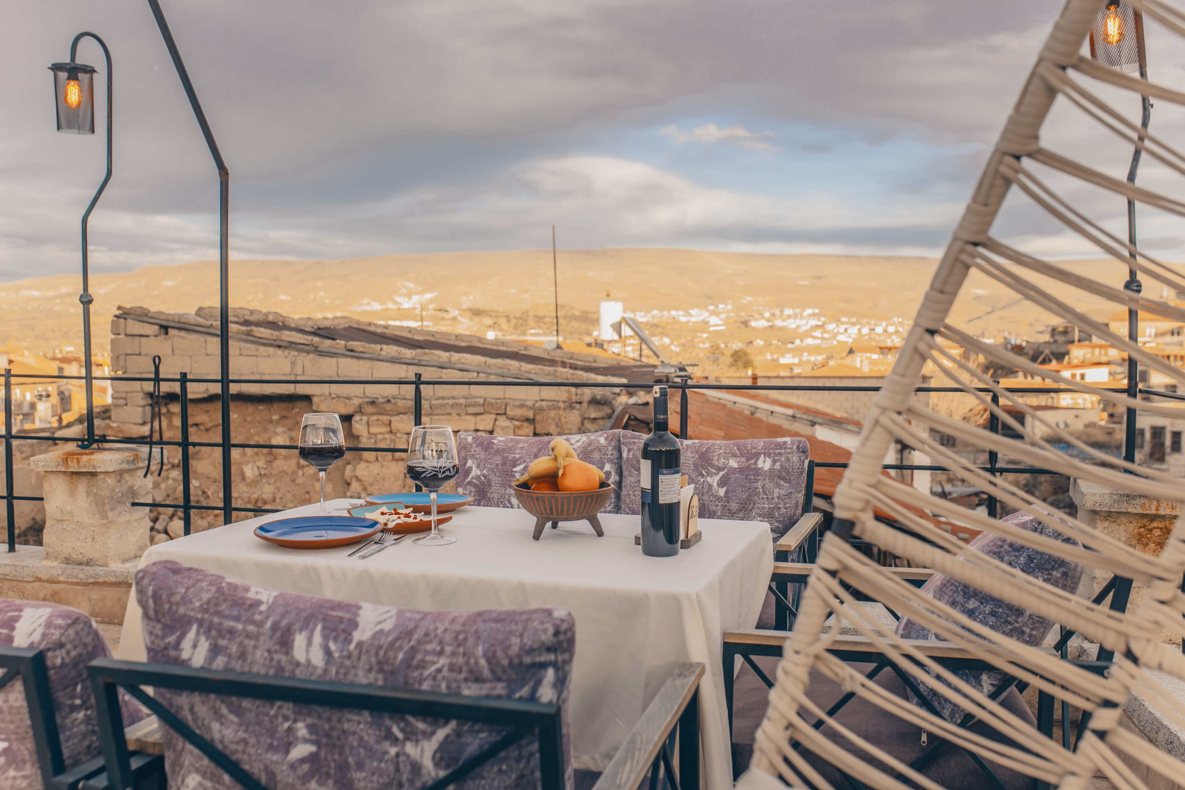 Mesa posta com copos de vinho, pratos e uma tigela de frutas, com vista para a paisagem urbana.
