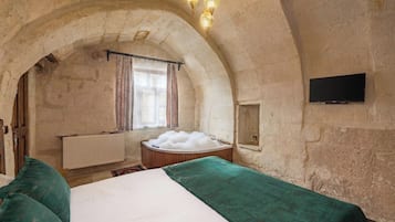 Quarto com arco de pedra, banheira, cama com roupa de cama verde, radiador e televisão fixada na parede.