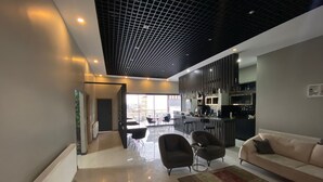 Uma sala de estar moderna com sofá, cadeiras e uma mesa de centro. Há uma cozinha com balcão e uma área de jantar com mesas e cadeiras.
