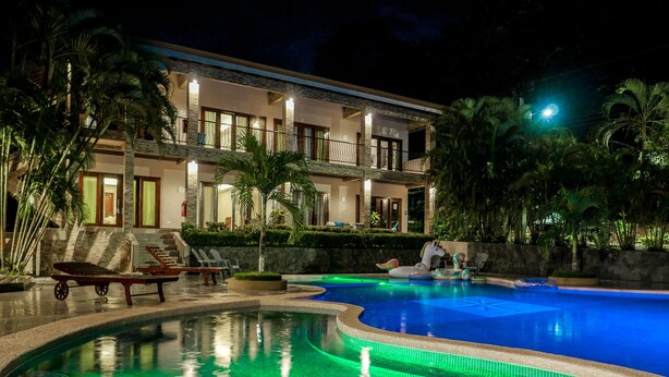 Uma piscina com design curvo, área de estar com cadeiras e um edifício com várias andares, varandas e iluminação.
