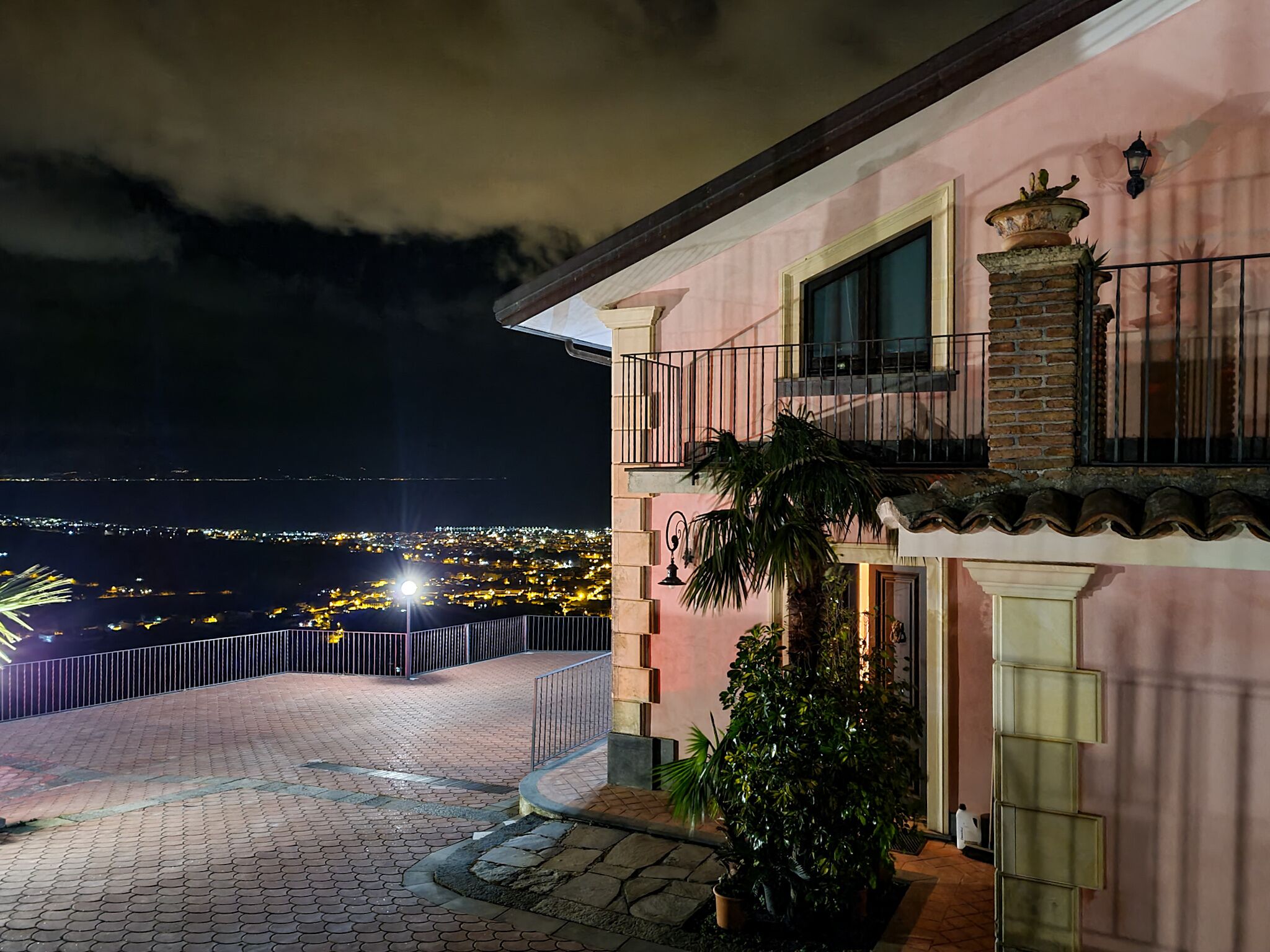 Um edifício de dois andares com varanda, entrada pavimentada e vista para o horizonte urbano à noite.