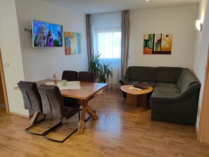 Uma sala de estar com uma mesa de jantar de madeira, duas cadeiras, um sofá verde e uma TV fixada na parede.