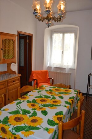 Uma sala de jantar com uma mesa coberta por um guardanapo de girassol, cadeiras de madeira, um candelabro, uma janela com cortinas e um radiador.
