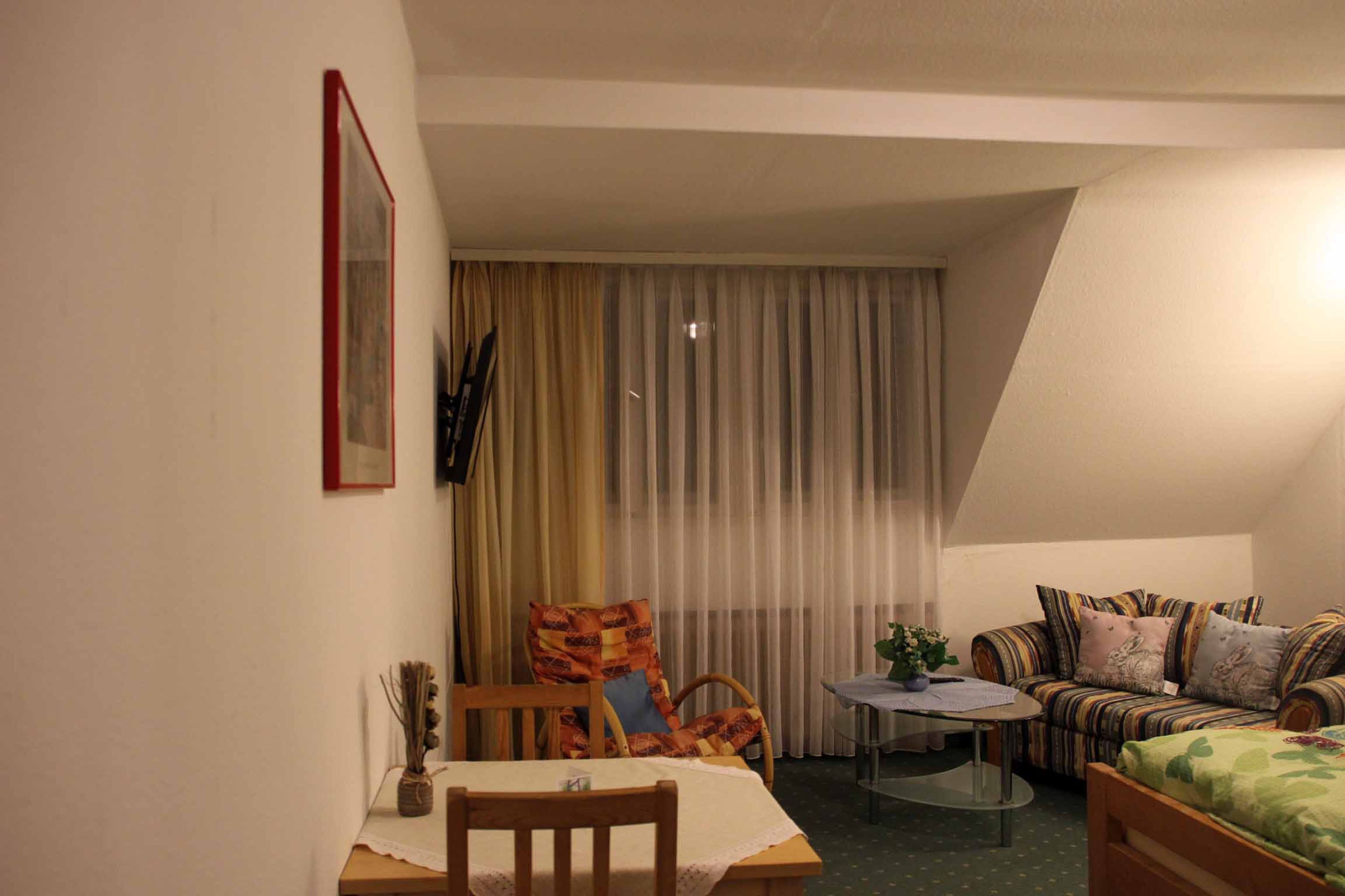 Quarto pequeno e acolhedor com uma mesa de jantar, um sofá e uma cama. Há uma janela com cortinas, um quadro na parede e uma pequena mesa com uma planta.