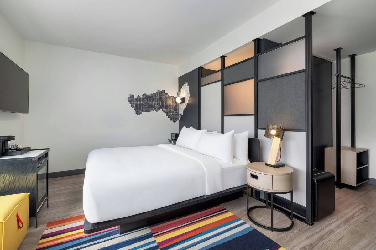Quarto de hotel moderno com uma cama grande, mesa de cabeceira com luminária, escrivaninha e uma obra de arte com um mapa na parede.