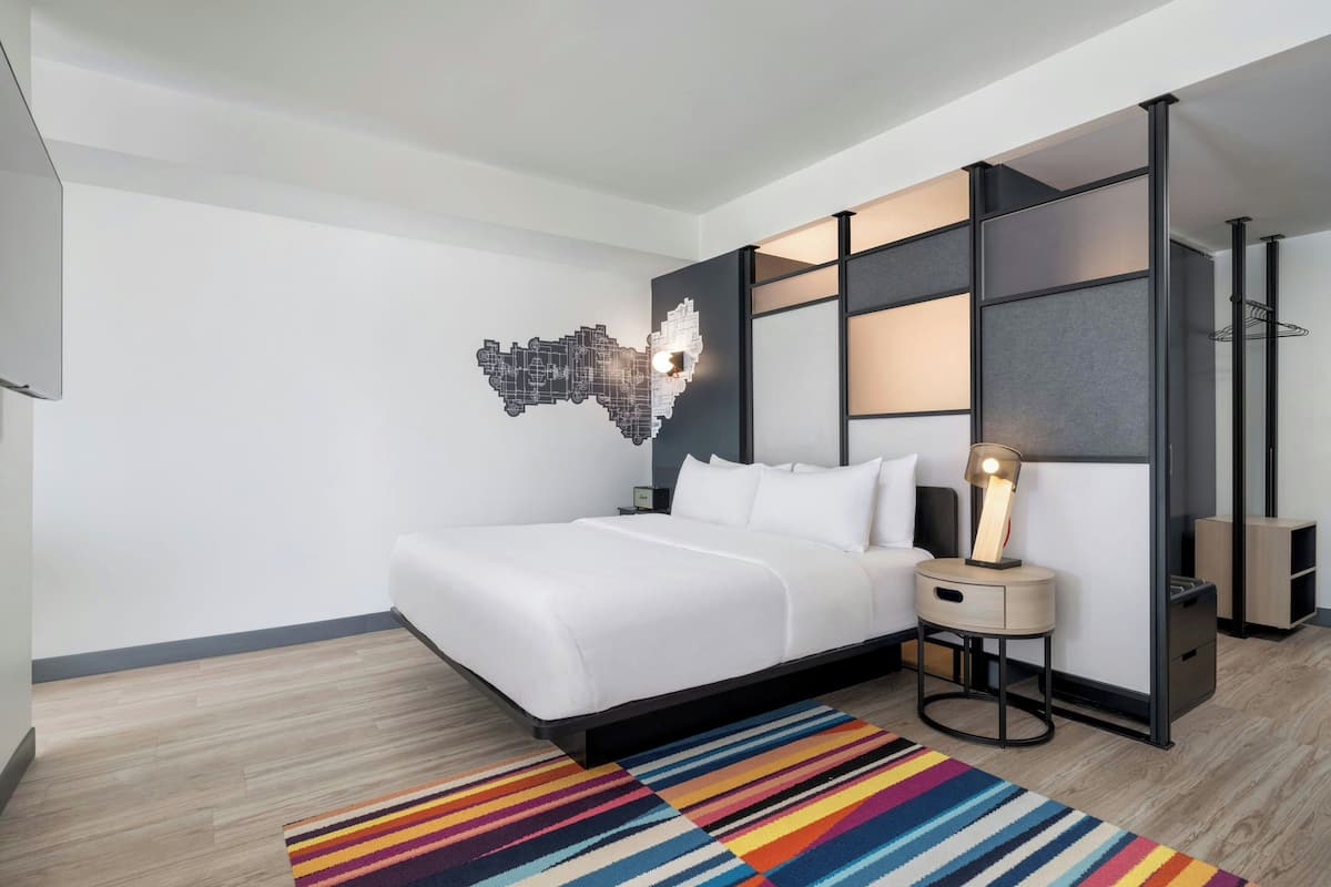 Quarto de hotel moderno com uma cama grande, um tapete listrado colorido, uma luminária fixada na parede e uma obra de arte com um mapa.