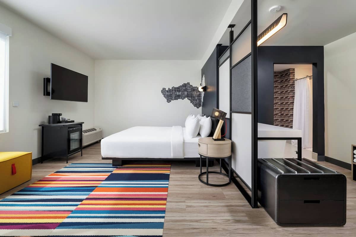 Quarto de hotel moderno com uma cama grande, televisão de tela plana, tapete listrado e colorido, mesinha lateral pequena e armário embutido.