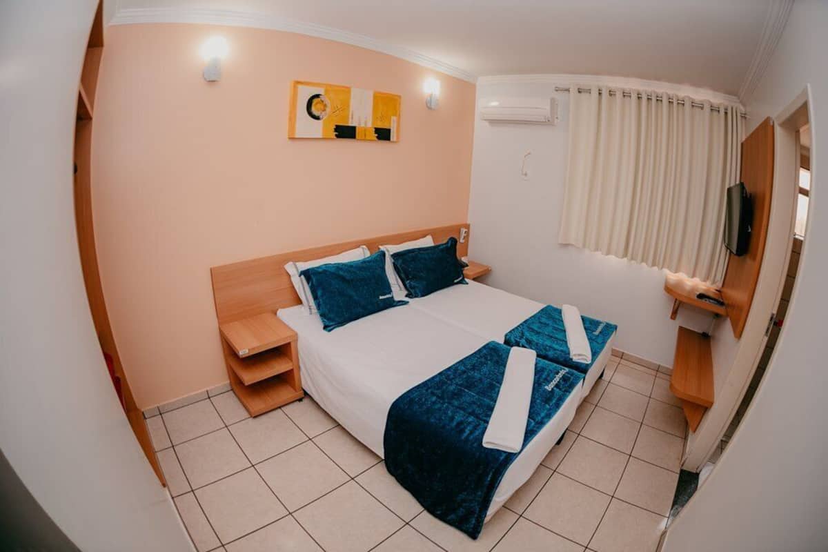 Quarto de hotel com cama, mesa de cabeceira, televisão na parede e janela com cortinas.