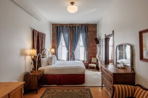 Quarto de hotel com uma cama grande, mesa de cabeceira de madeira, cômoda, cadeira e espelho.