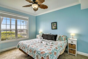 Quarto com cama, duas mesinhas de cabeceira, ventoinha no teto, janela com persianas e vista para o verde lá fora.