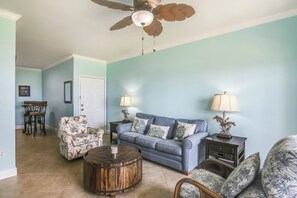 Uma sala de estar com um sofá azul, poltronas estampadas, uma mesa de centro de madeira e um ventilador de teto.