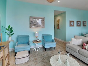Uma sala de estar com dois poltronas azuis, um sofá cinza, uma mesa redonda pequena com uma luminária, e um quadro emoldurado na parede.