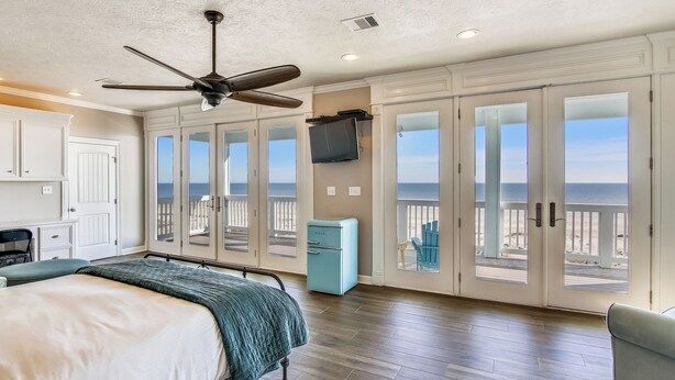 Quarto com cama, ventilador de teto, televisão e vista para o mar através de grandes portas deslizantes.