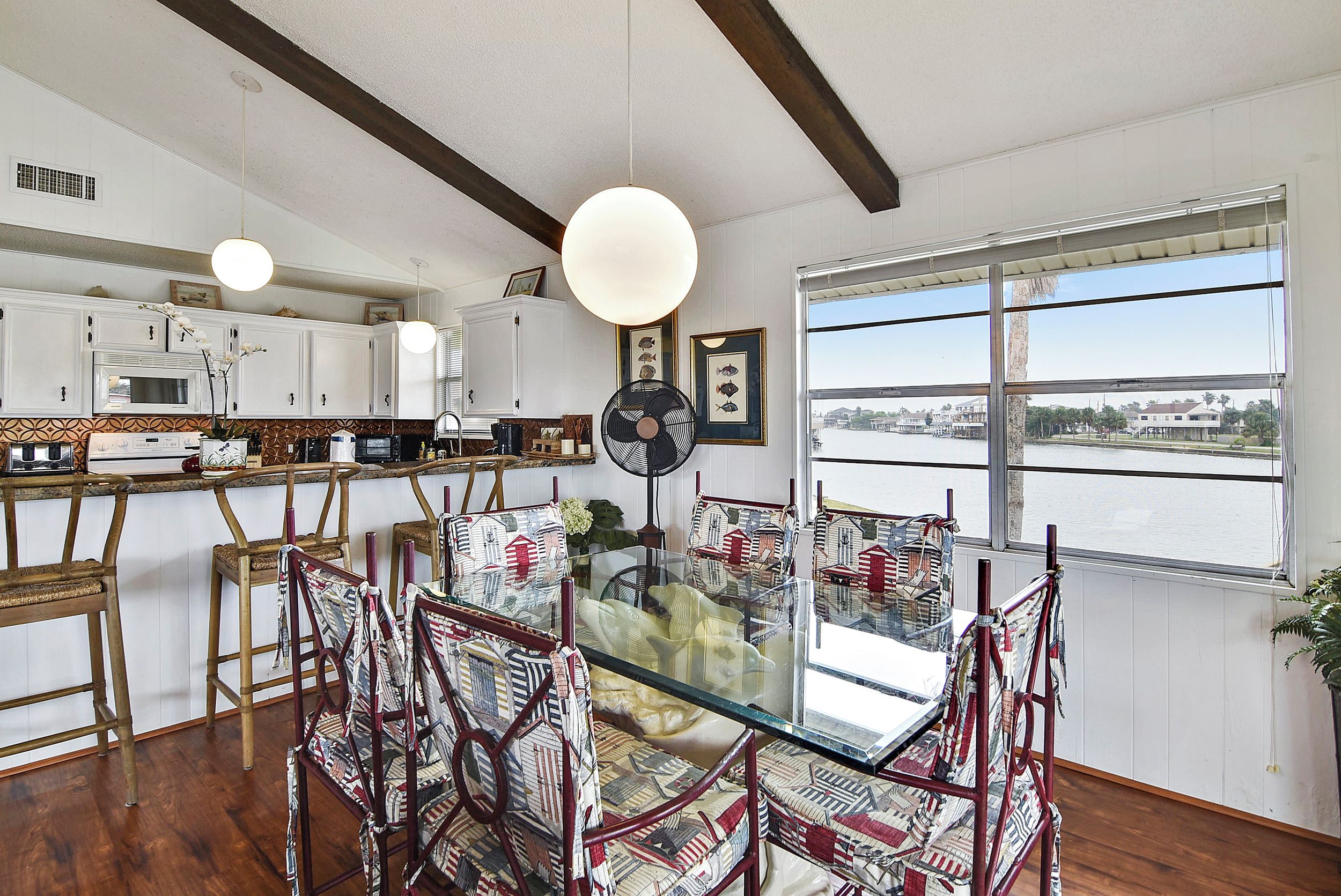 Uma sala de jantar com mesa de vidro cercada por cadeiras estampadas, armários brancos e uma janela grande com vista para um corpo d'água.