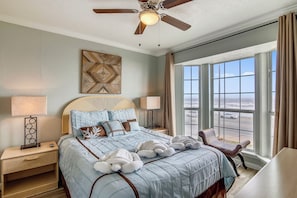 Quarto com cama grande, duas mesas de cabeceira, ventilador de teto, vista para o oceano e janela com cortinas.