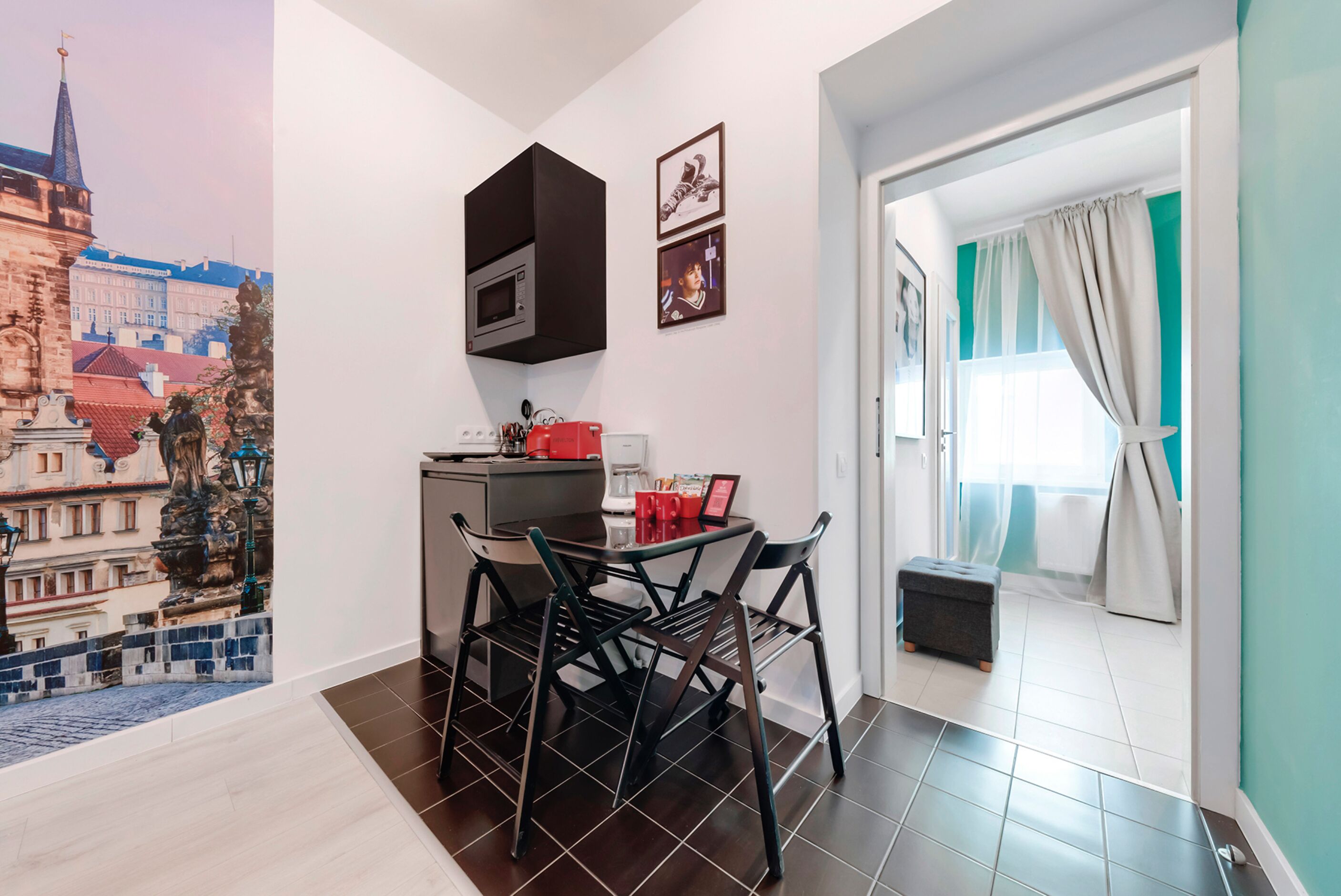 Quarto compacto com cozinha americana, área de jantar com mesa e cadeiras, um pequeno banquinho e vista para a cidade através de uma janela.