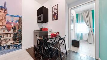 Quarto compacto com cozinha americana, área de jantar com mesa e cadeiras, um pequeno banquinho e vista para a cidade através de uma janela.