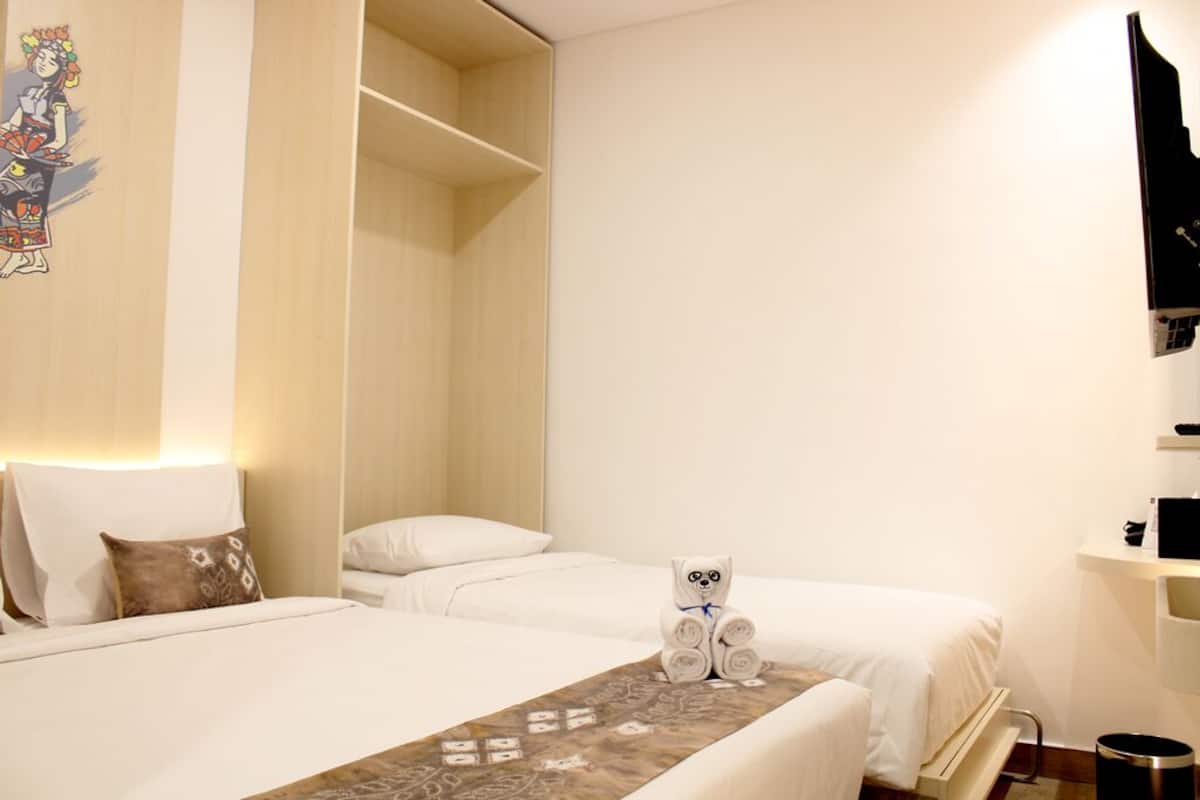 Quarto de hotel com duas camas, um guarda-roupa, uma mesa pequena com um urso de pelúcia e uma televisão fixada na parede.