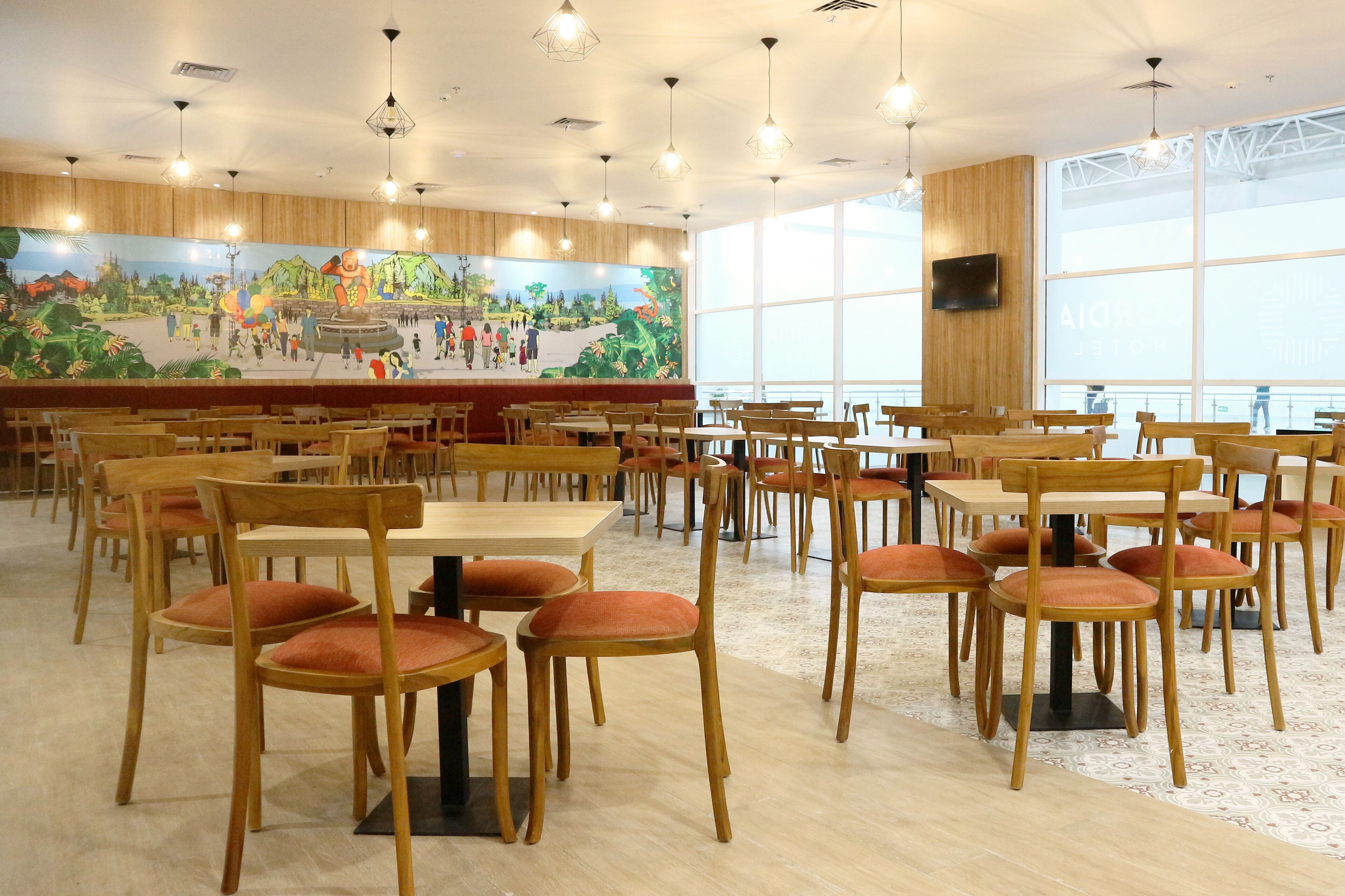 Uma sala de jantar espaçosa com mesas e cadeiras de madeira, um mural na parede e grandes janelas.