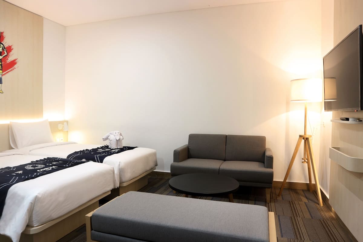 Quarto de hotel com duas camas, um sofá, uma mesa redonda pequena, uma televisão e um abajur.