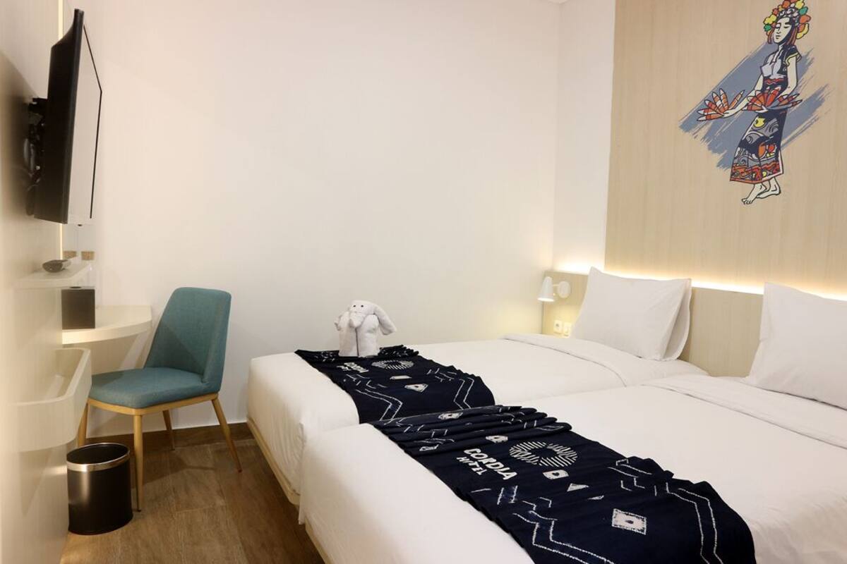Quarto de hotel com duas camas, uma escrivaninha, uma cadeira, um cesto de lixo, uma televisão e um objeto decorativo na parede.