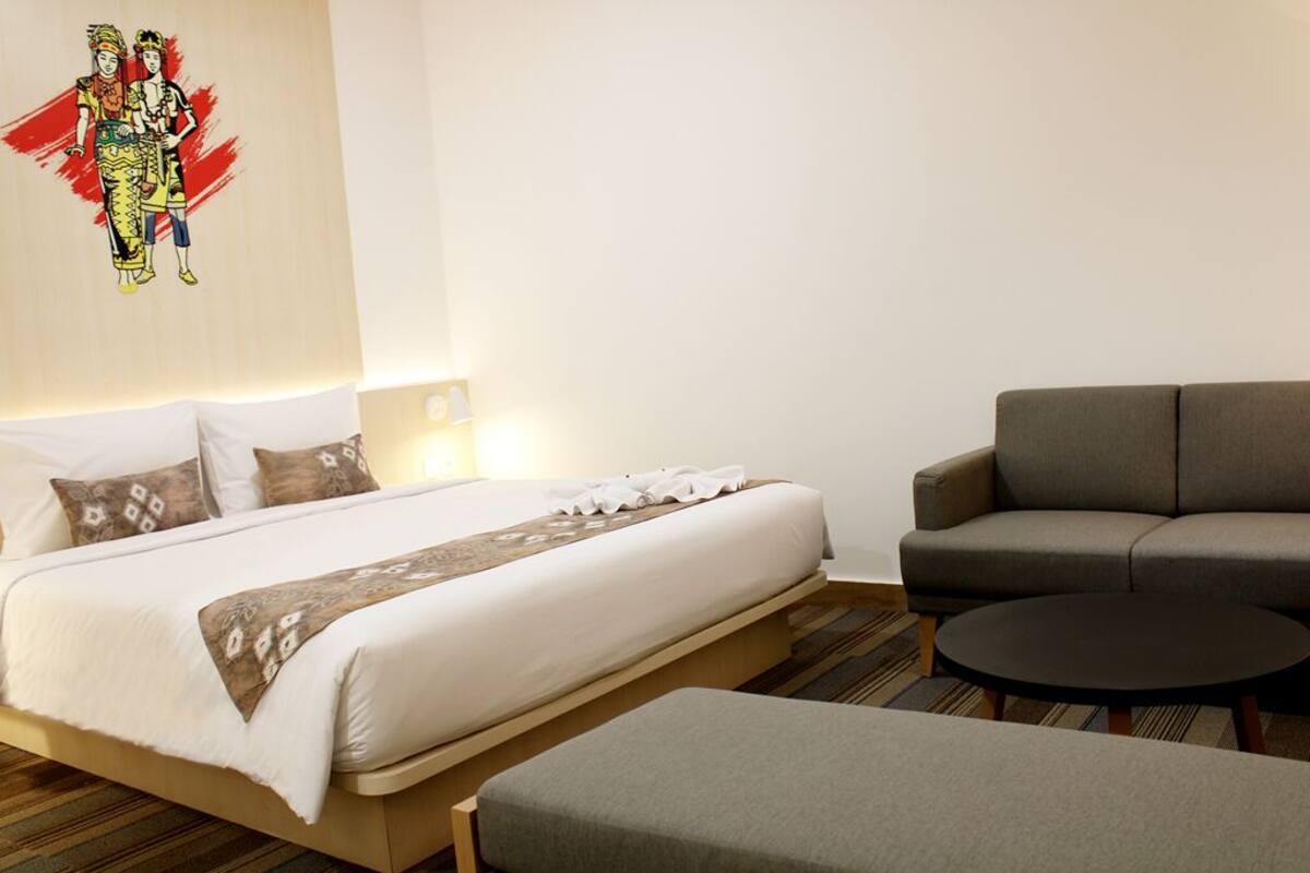 Quarto de hotel com uma cama, um sofá cinzento, uma pequena mesa redonda e uma parede com um design abstrato em vermelho.