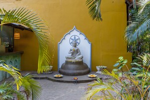 Estátua de uma figura sentada em um nicho com arco decorativo, rodeada por plantas e flores.