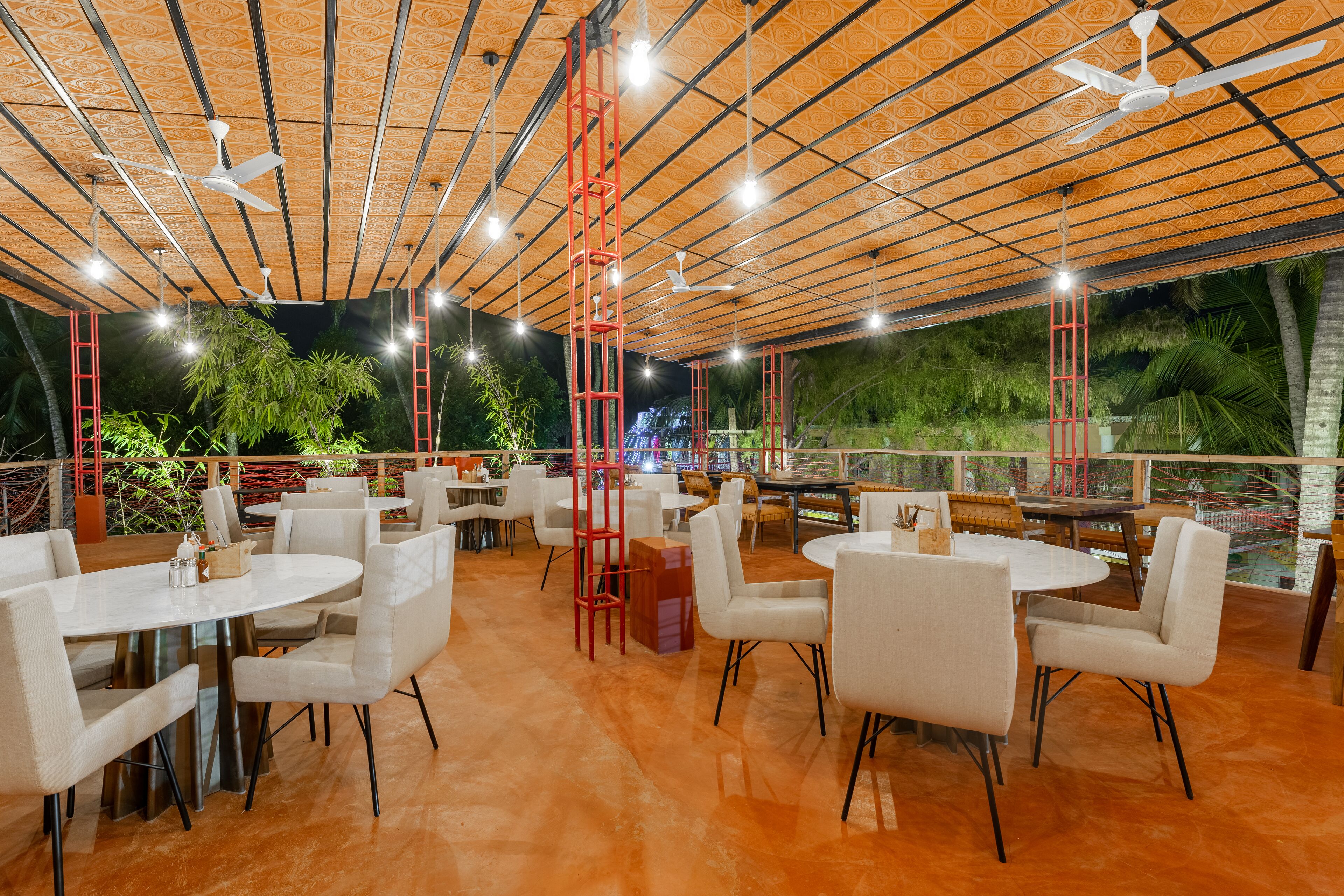Restaurante ao ar livre com mesas redondas e cadeiras brancas, teto de madeira com padrão quadriculado e estruturas metálicas vermelhas.