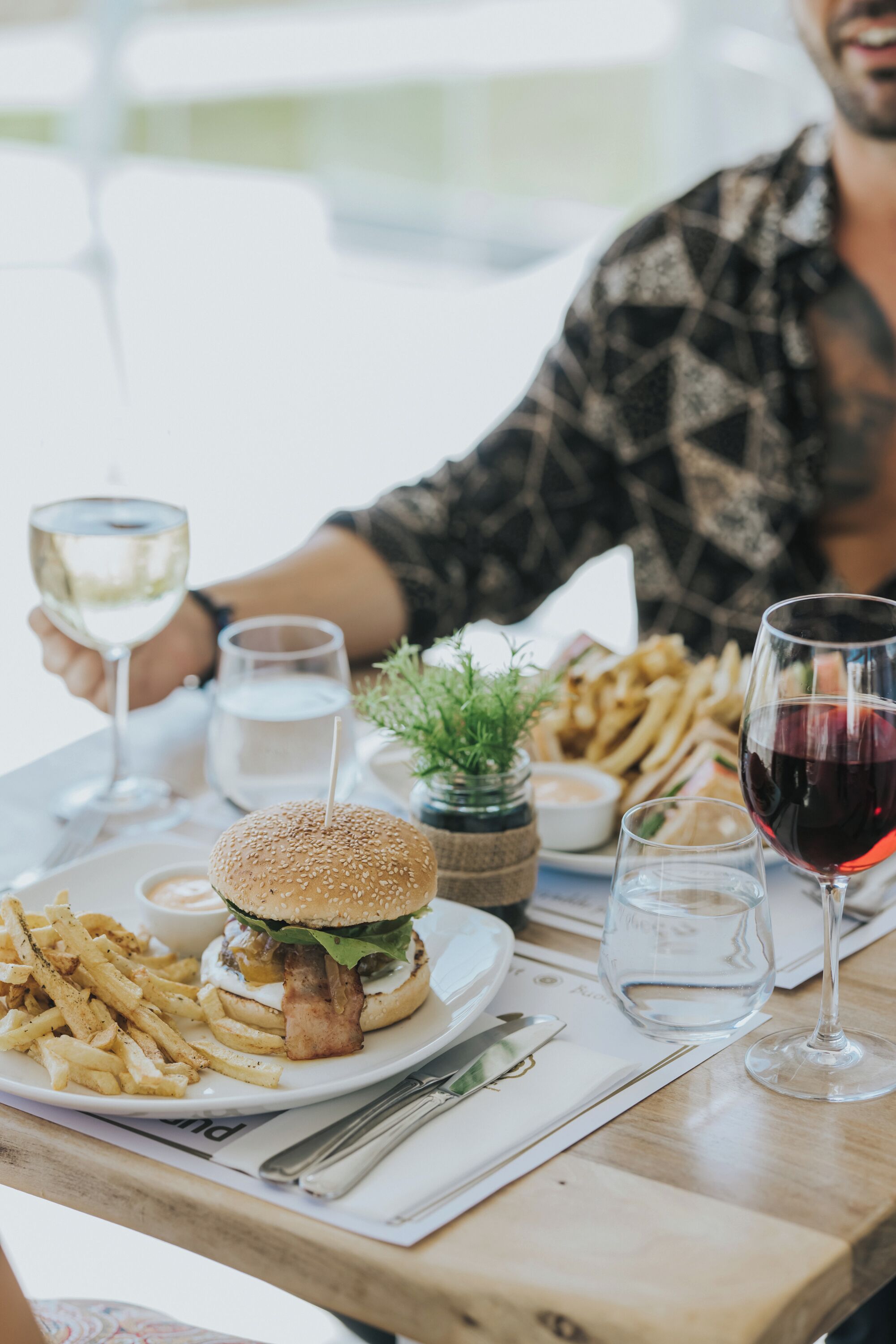 Uma mesa de jantar com um hambúrguer, batatas fritas e duas taças de vinho.