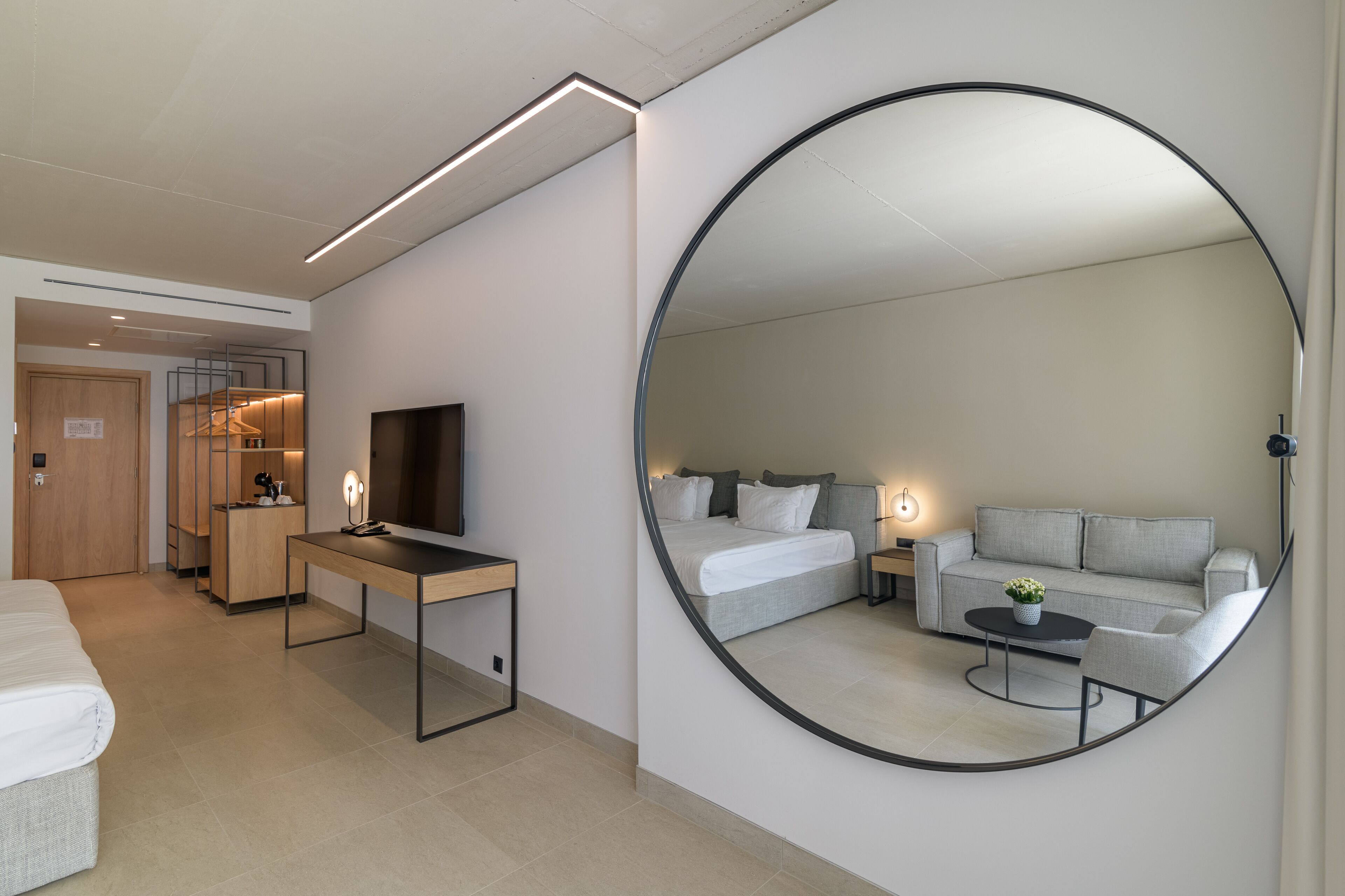 Quarto de hotel moderno com um espelho redondo, uma cama, um sofá, uma escrivaninha e uma pequena mesa com uma planta.